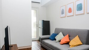 Uma sala de estar com um sofá cinzento, almofadas coloridas, uma televisão de tela plana e uma geladeira.