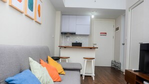 Sala de estar com sofá cinza, almofadas coloridas, televisão e área de cozinha com armários brancos e bancada de madeira.