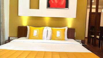 Quarto de hotel com uma cama, duas almofadas amarelas com a marca 'bloom hotels' e um quadro abstrato grande, além de uma cabeceira de madeira.