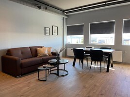 Uma sala de estar moderna com um sofá de couro marrom, uma mesa de centro redonda e uma área de jantar com mesa e quatro cadeiras. Há três quadros na parede e janelas amplas com persianas.