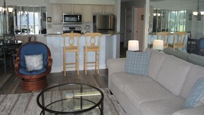Uma sala de estar moderna com um sofá, uma mesa de centro de vidro, uma poltrona azul, cadeiras de madeira e uma cozinha ao fundo.