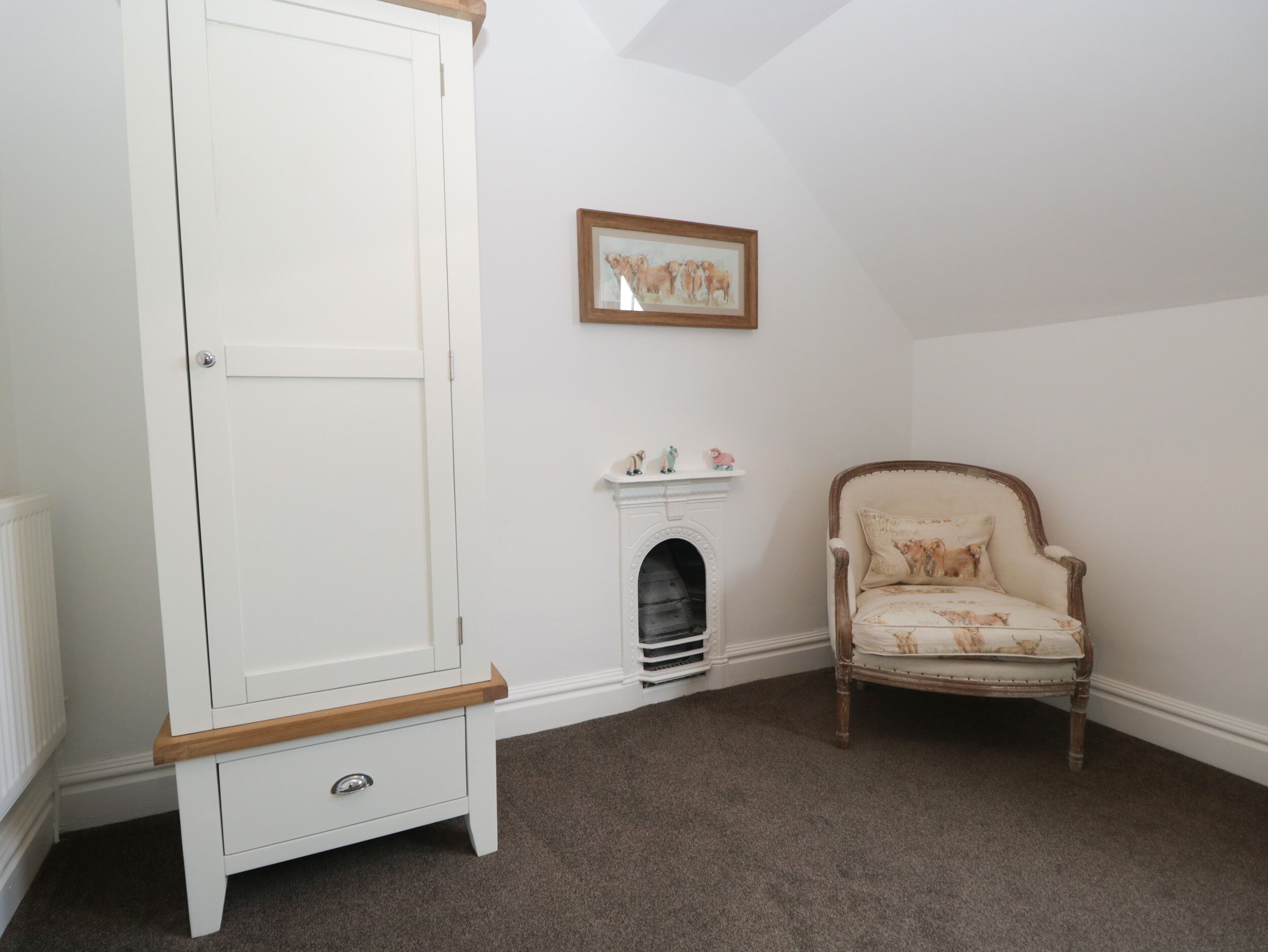 Um quarto com um guarda-roupa branco, uma pequena lareira decorada com figuras e uma cadeira com almofada florida.