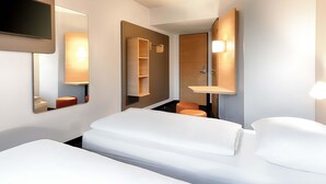 Quarto de hotel moderno com duas camas, um espelho grande, uma prateleira de madeira e um pequeno espaço de estar.