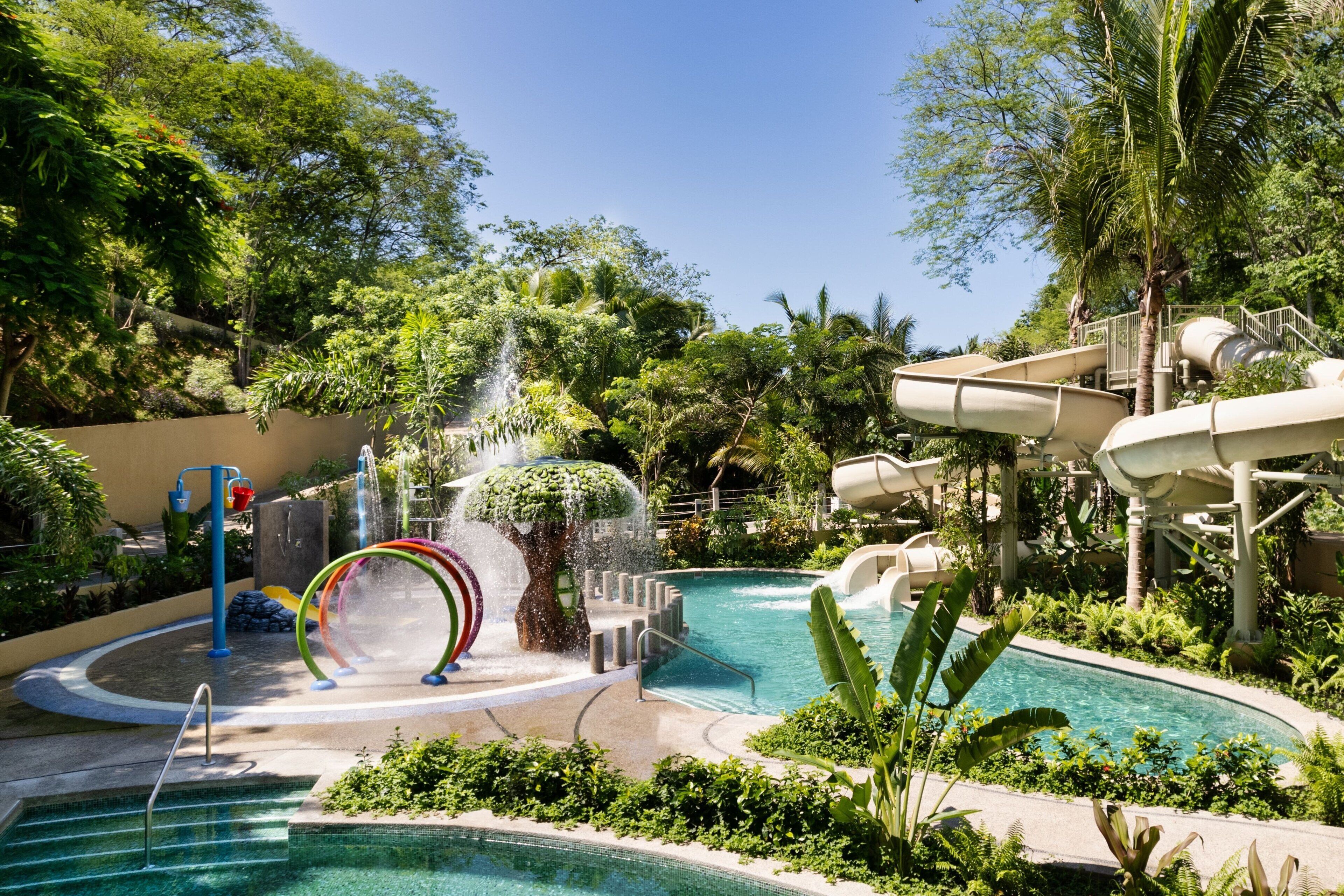 Uma área de piscina com toboáguas, um atrativo aquático circular e vegetação exuberante ao redor.