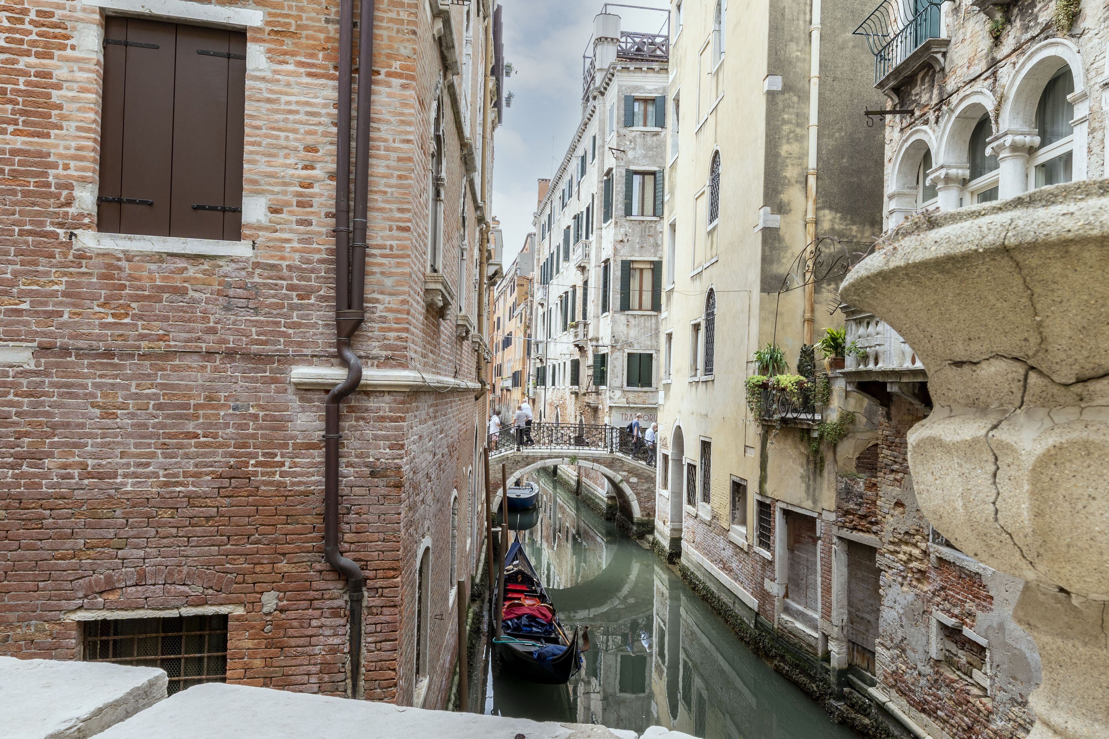 Um canal estreito numa cidade europeia histórica, com uma gôndola, edifícios de tijolo e uma estrutura de pedra.