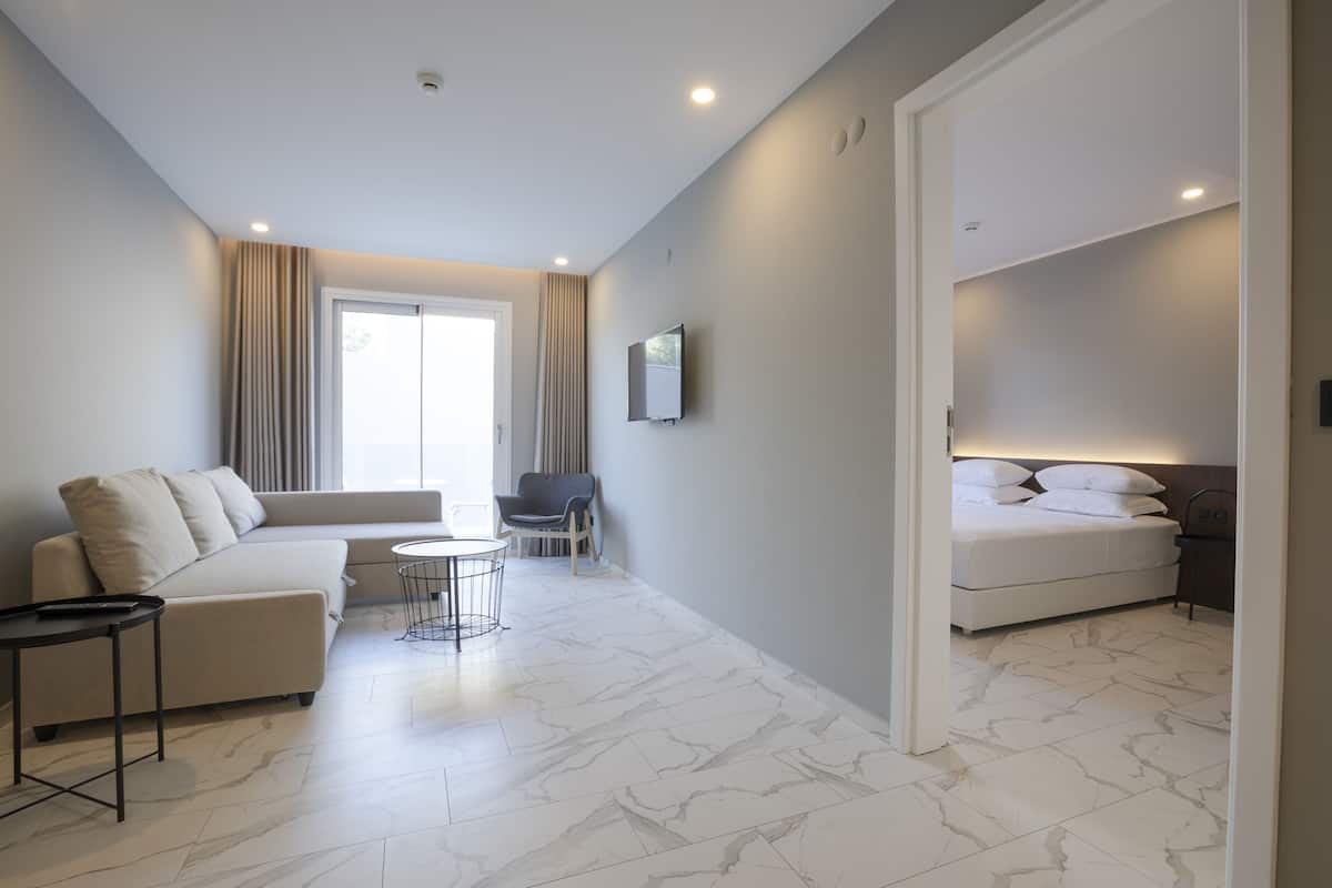 Quarto de hotel moderno com sofá, mesa redonda pequena, cadeira, cama e televisão presa na parede.