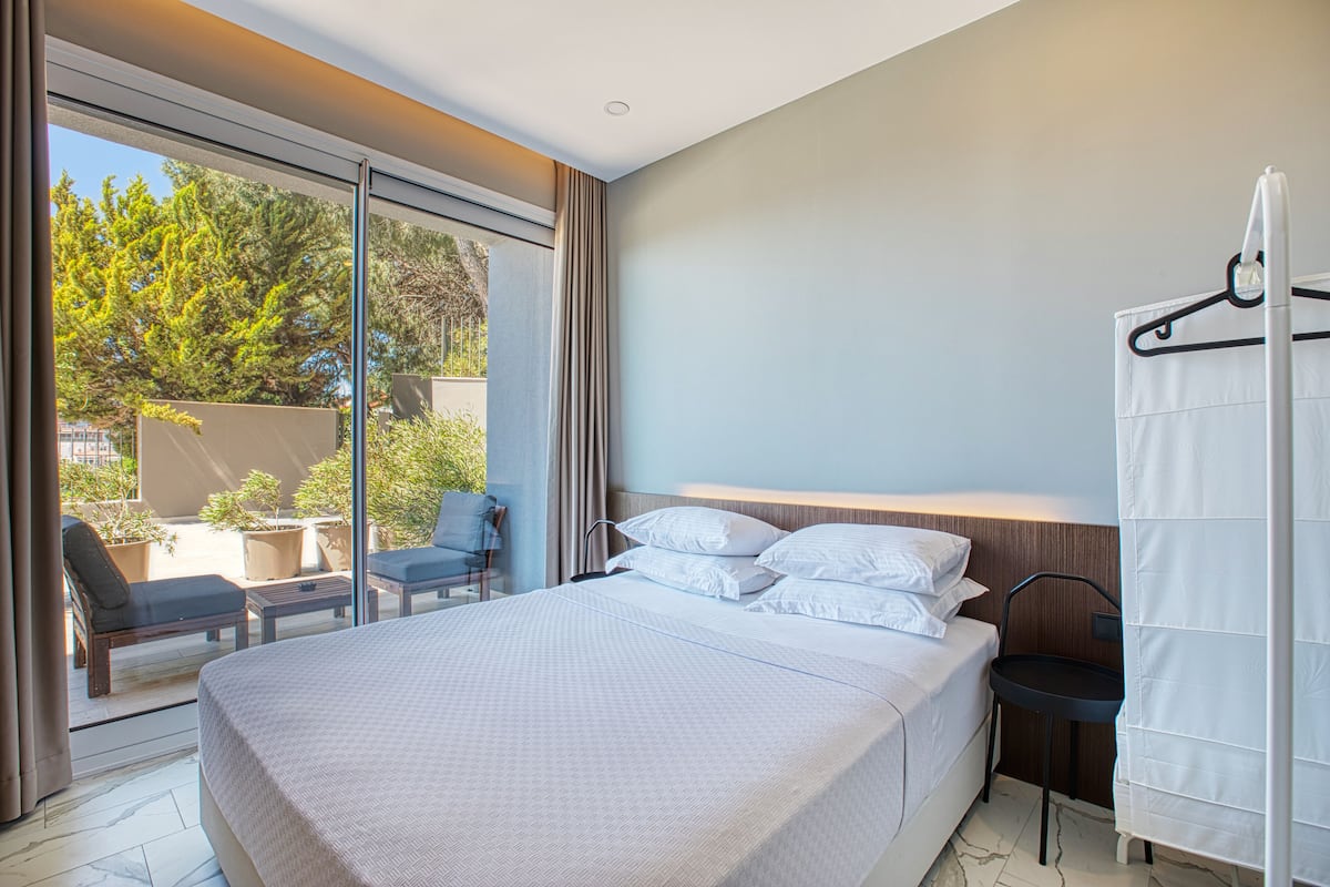 Quarto de hotel moderno com uma cama grande, uma mesinha, uma cadeira e vista para o exterior.