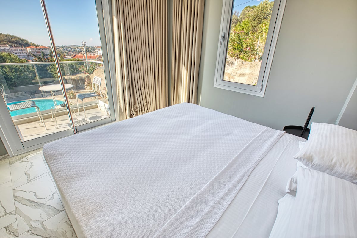 Quarto de hotel com uma cama grande, vista para uma piscina e varanda com mesa e cadeiras.