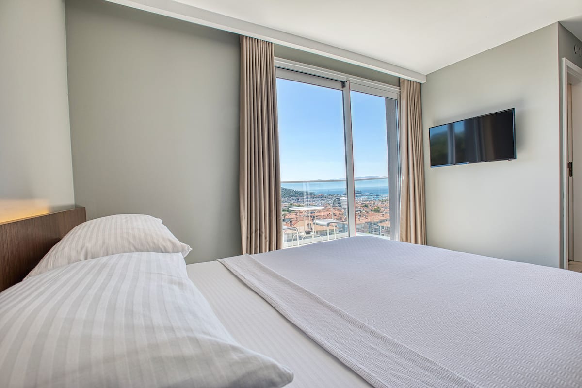 Quarto de hotel com uma cama grande, televisão fixada na parede e janela com vista para a cidade.