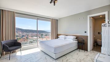 Quarto de hotel com uma cama grande, uma cadeira e vista para a paisagem urbana através de uma janela ampla.