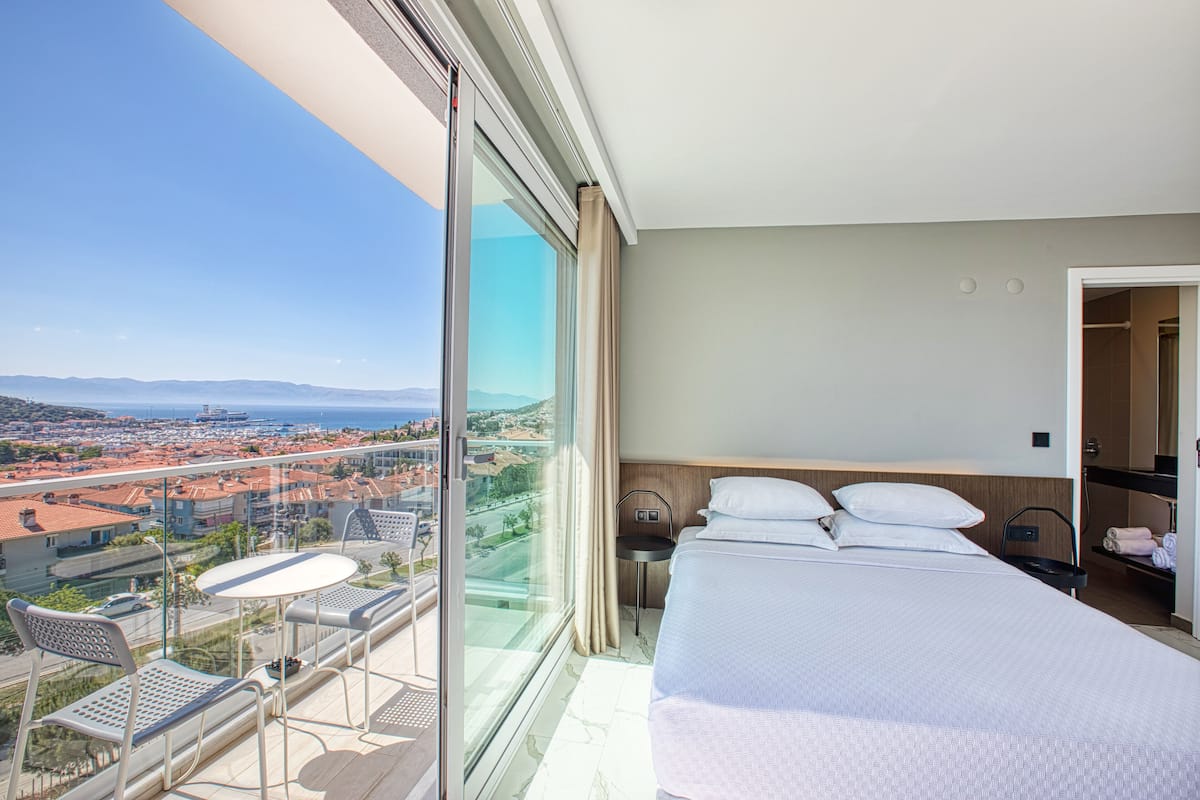 Quarto de hotel com cama grande, varanda com mesa e cadeiras, e vista para a cidade.