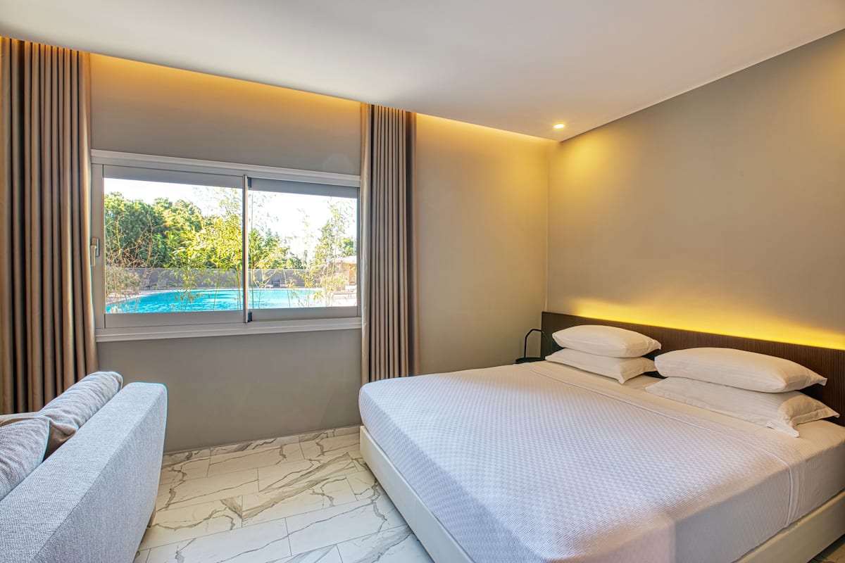 Quarto de hotel com uma cama, um sofá, uma janela com vista para uma piscina e piso de mármore.