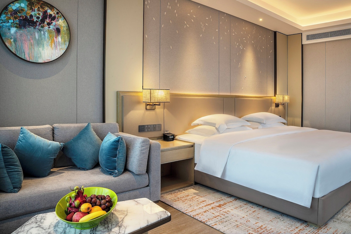 Quarto de hotel com cama, sofá, mesa de cabeceira, abajur e uma obra de arte na parede.