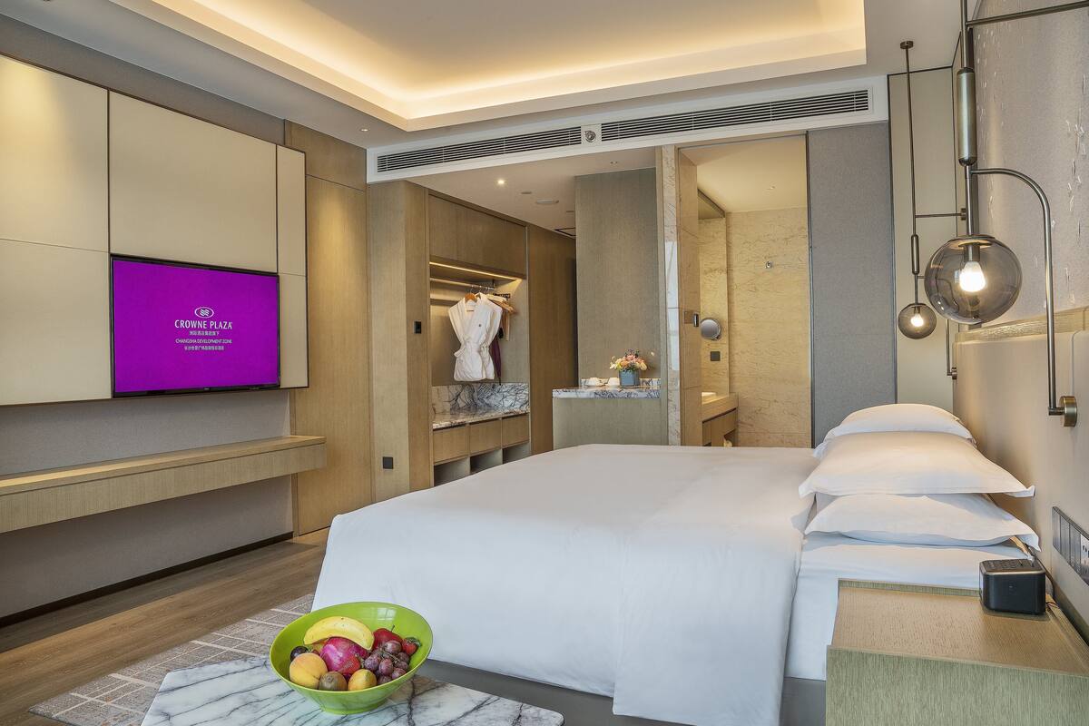 Quarto de hotel moderno com uma cama grande, televisão na parede, banheiro com espelho e uma cesta de frutas no chão.