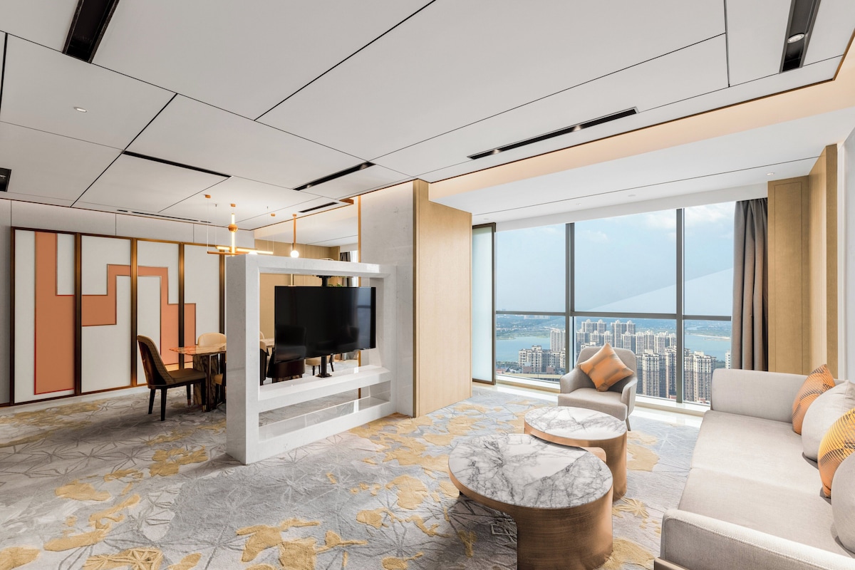 Quarto de hotel moderno com televisão grande, área de estar e vista para a cidade.