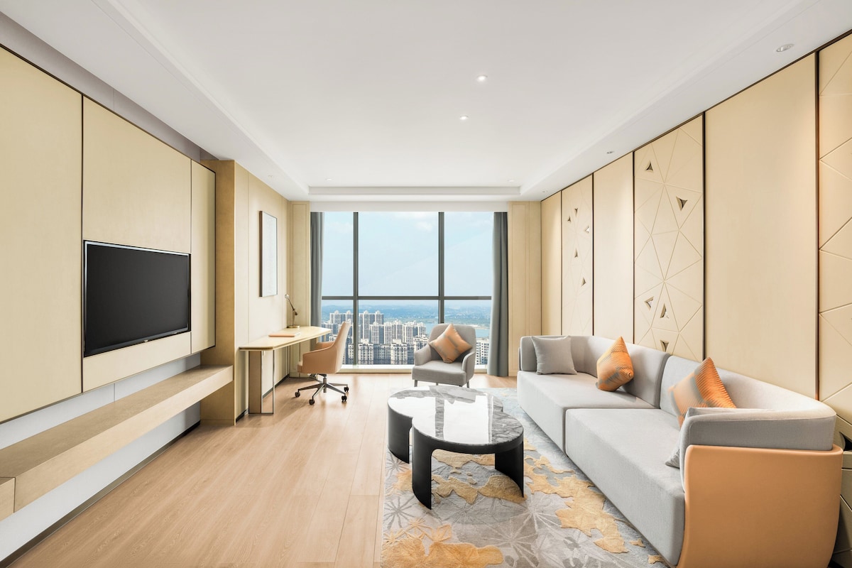 Uma sala de estar moderna com uma televisão grande, assentos confortáveis, uma escrivaninha e vista para um horizonte urbano através de grandes janelas.
