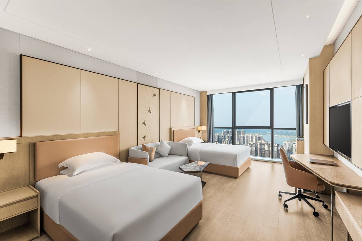 Quarto de hotel moderno com duas camas, uma escrivaninha e uma janela ampla com vista para a cidade.