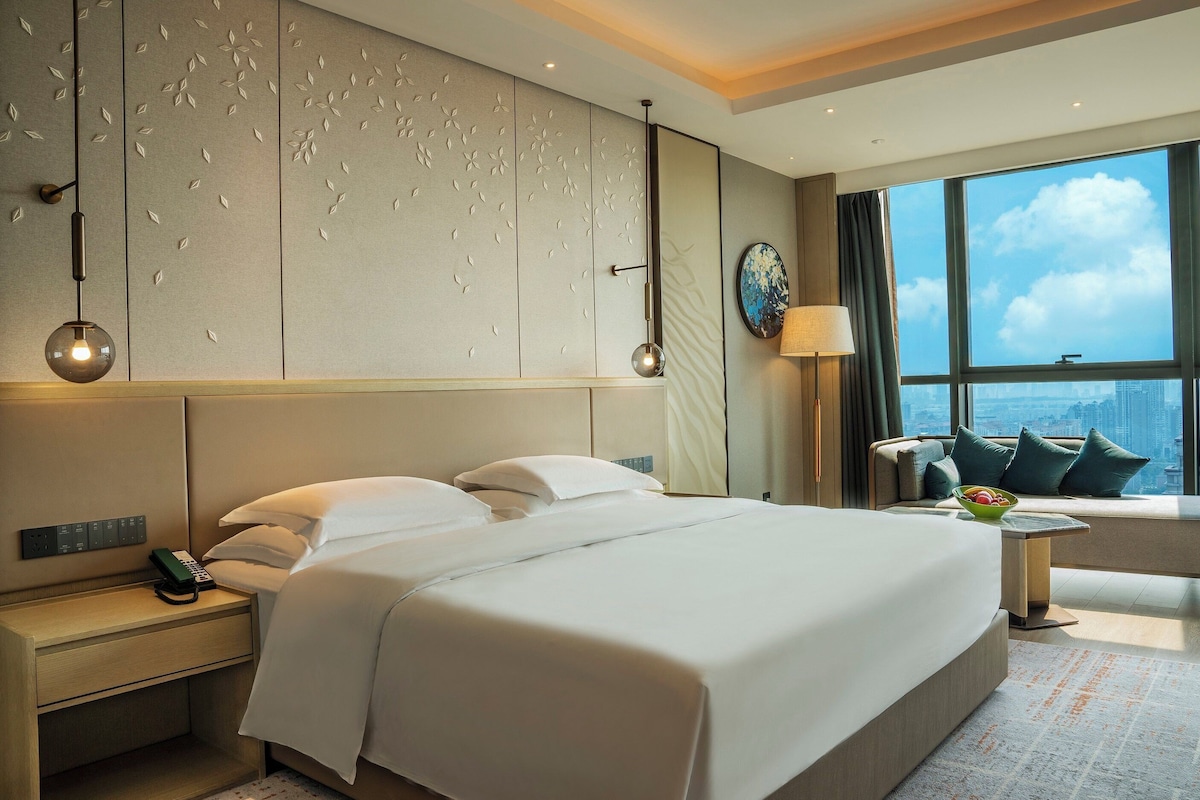 Quarto de hotel moderno com uma cama grande, mesa de cabeceira, luminária e vista da cidade por amplas janelas.