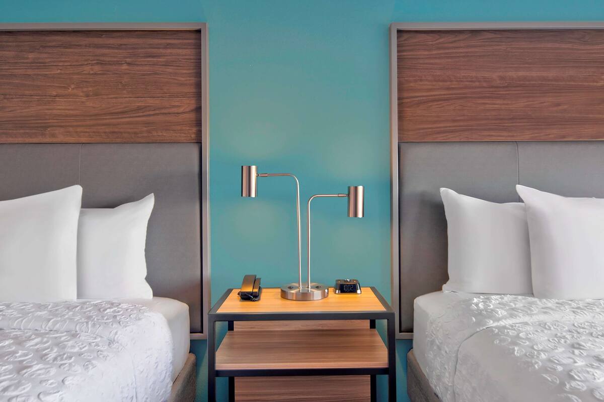 Quarto de hotel com duas camas, uma mesinha de cabeceira de madeira, um relógio digital e uma luminária fixada na parede.