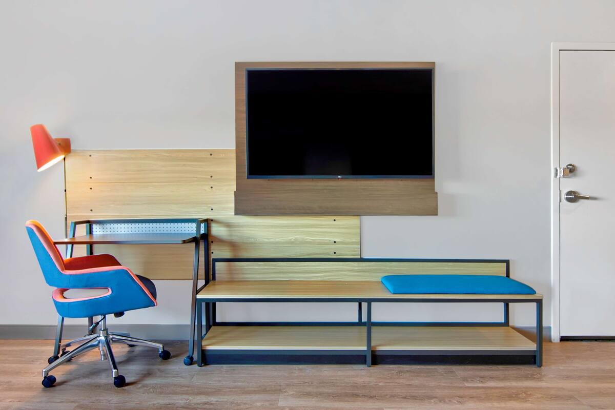 Quarto moderno com escrivaninha de madeira, cadeira azul e laranja, um banco e TV na parede.