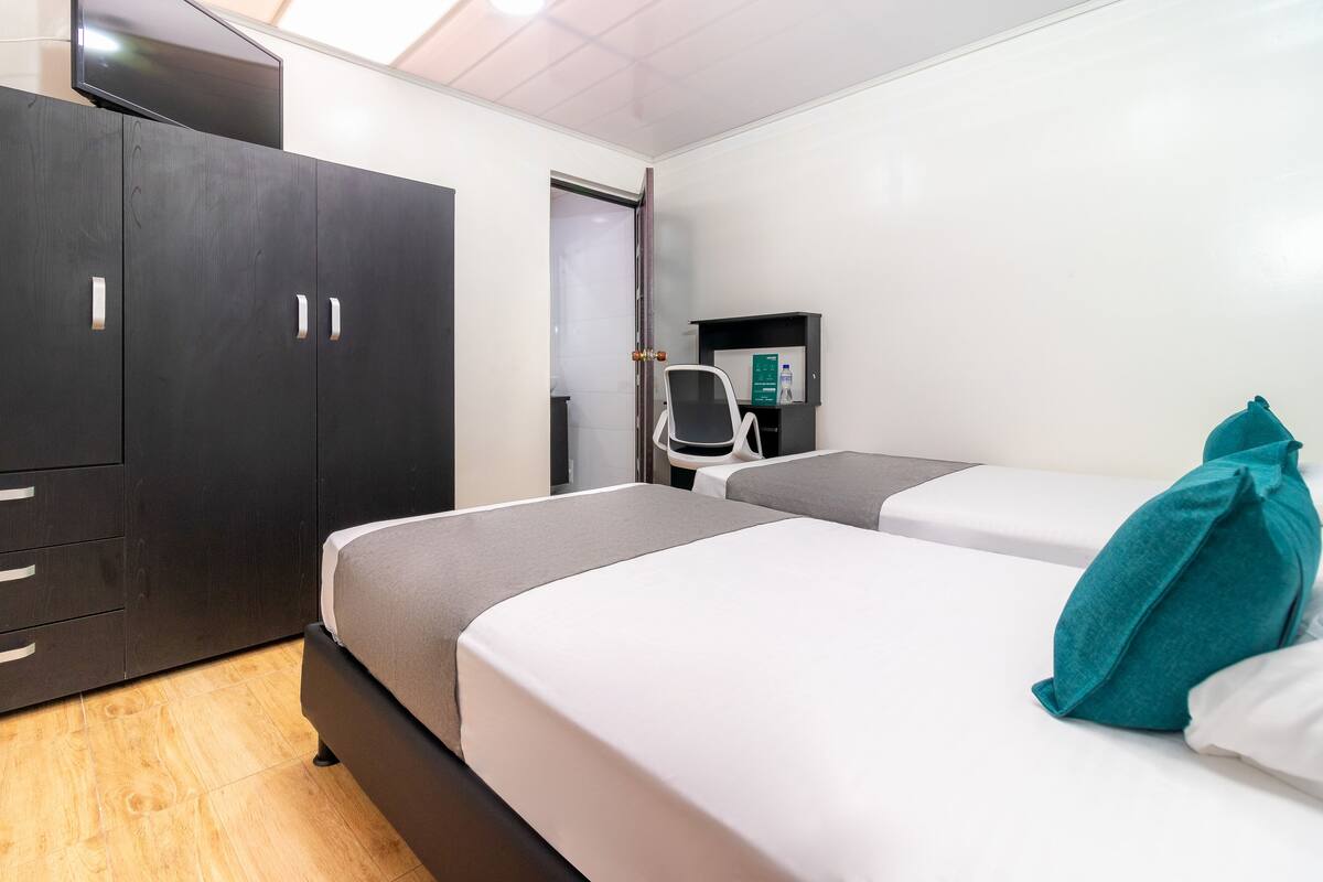 Quarto de hotel moderno com um guarda-roupa espaçoso, uma cama com almofada azul-turquesa e uma escrivaninha com cadeira.