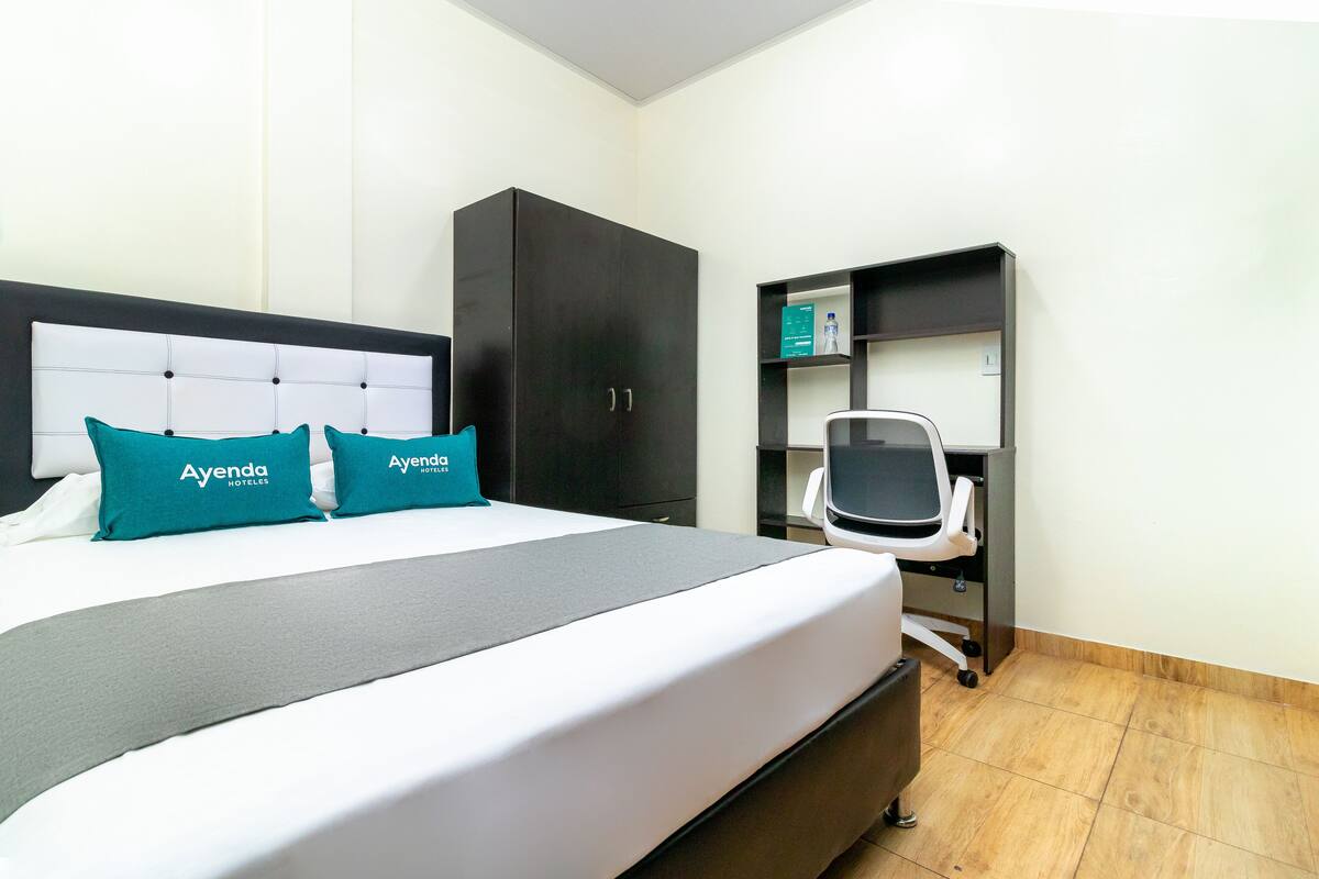 Quarto de hotel com cama, dois travesseiros verde-água, armário escuro, escrivaninha com cadeira e prateleira com garrafa de água.