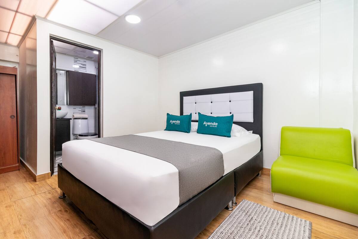 Quarto de hotel com uma cama, uma poltrona verde, banheiro com espelho e pia, e piso de madeira.
