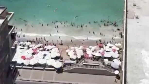 Uma praia com águas turquesa cristalinas e vista para o oceano. Há pessoas na praia e guarda-sóis montados.