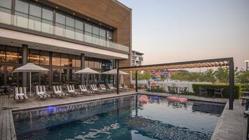 Piscina no terraço com espreguiçadeiras e guarda-sóis, emoldurada por um deck de madeira e um edifício moderno com grandes janelas.