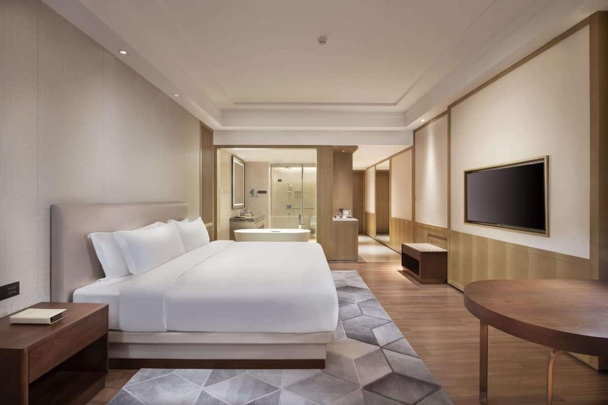 Quarto de hotel moderno com uma cama grande, uma mesa de cabeceira, uma televisão e um banheiro visível por uma porta aberta.