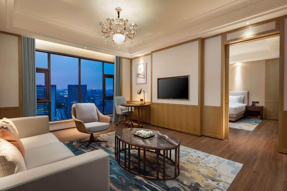 Quarto de hotel moderno com uma janela ampla que oferece vista para a cidade, um espaço de estar confortável, uma televisão de tela plana e uma cama aconchegante.