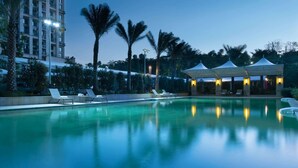 Piscina com espreguiçadeiras e área coberta com iluminação, cercada por palmeiras e um edifício ao fundo.