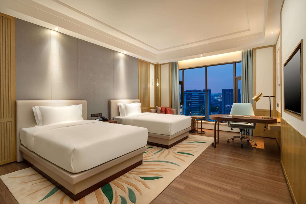 Quarto de hotel com duas camas, uma escrivaninha, uma cadeira e uma janela grande com vista para a cidade.