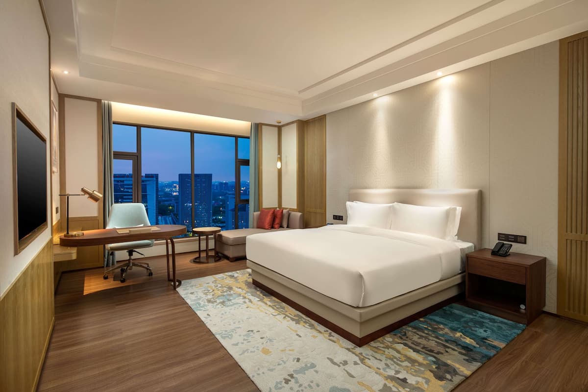 Quarto de hotel moderno com uma cama grande, uma escrivaninha, uma cadeira e vista para a cidade.