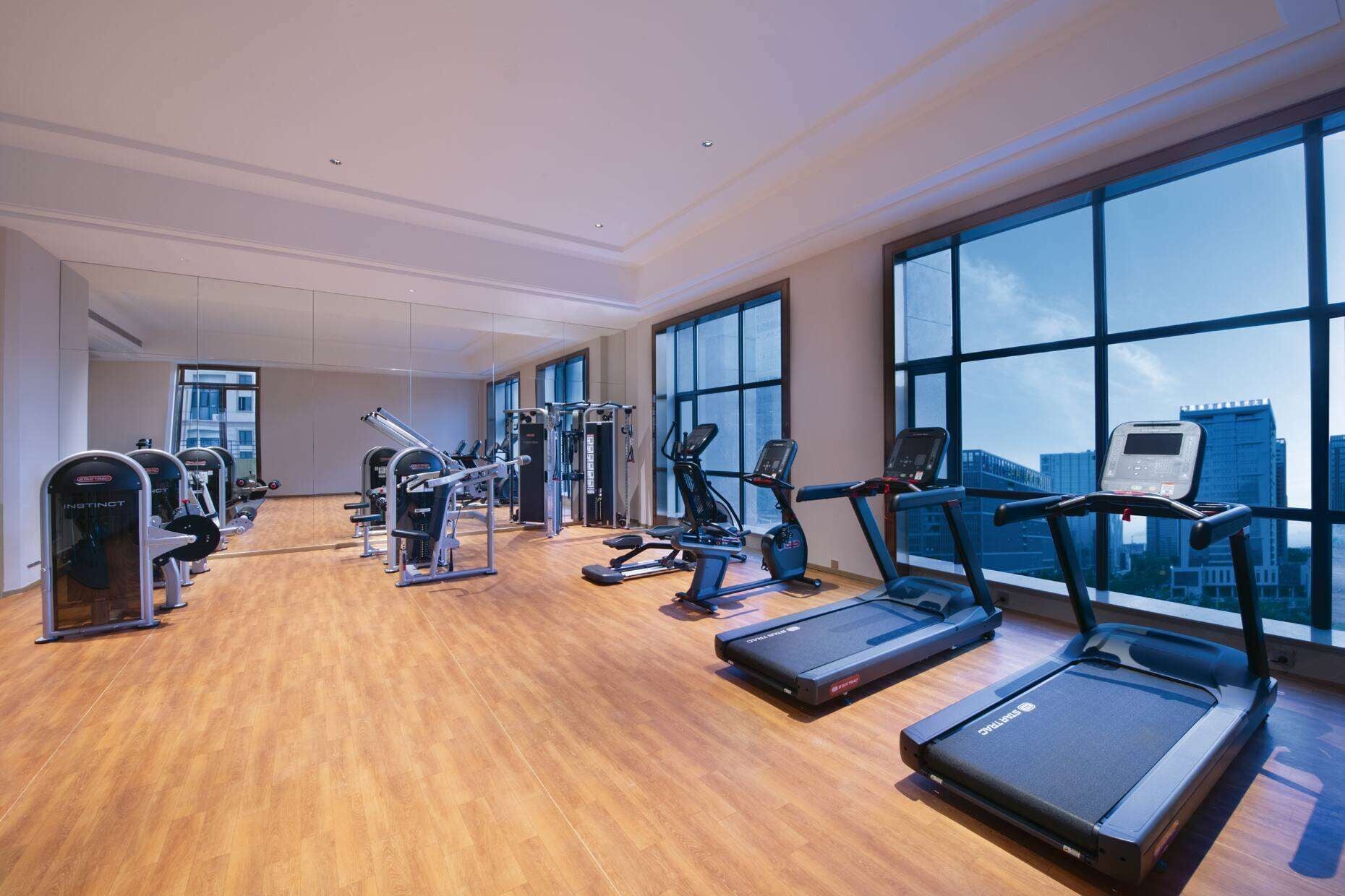 Uma academia espaçosa com diversos equipamentos de exercício, amplas janelas com vista para a cidade e piso de madeira.