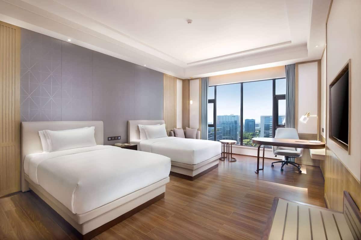 Quarto de hotel moderno com duas camas, uma escrivaninha, uma cadeira e uma janela grande com vista para a cidade.