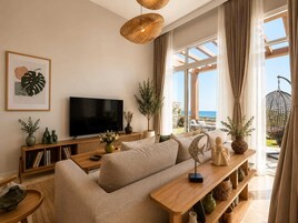Sala de estar moderna com televisão de tela plana grande, sofá, mesa de centro de madeira e vista para o oceano através de grandes janelas.