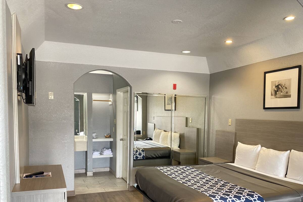 Quarto de hotel moderno com uma cama grande, uma televisão fixada na parede e um banheiro visível através de uma porta em arco.