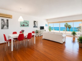 Uma sala de estar moderna com um sofá branco, cadeiras vermelhas, uma mesa de jantar e uma grande janela com vista para o mar.
