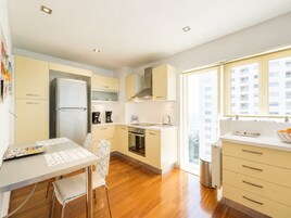 Uma cozinha moderna com geladeira, forno e cafeteira. Há uma mesa de jantar com cadeiras e uma janela com vista para edifícios.