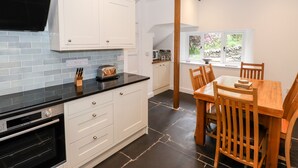 Uma cozinha com armários brancos, bancada preta e forno Bosch. Há uma área de jantar com mesa e cadeiras de madeira, e uma janela com vista para o verde.