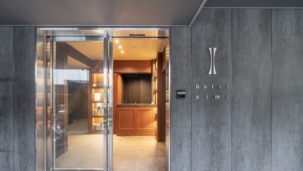 Entrada de hotel com design moderno, com porta de vidro, balcão de recepção e área de cozinha visível.