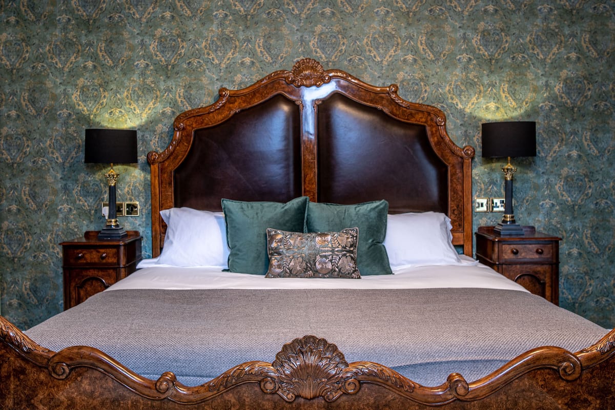 Uma cama grande com cabeceira de madeira, duas mesas de cabeceira e um papel de parede estampado.