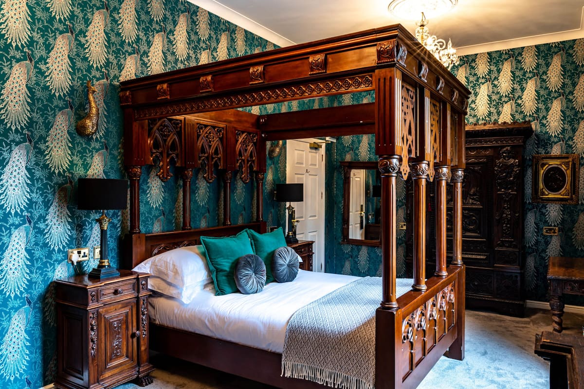 Uma cama com dossel de estrutura de madeira e detalhes entalhados, um criado-mudo com abajur e um espelho na parede.