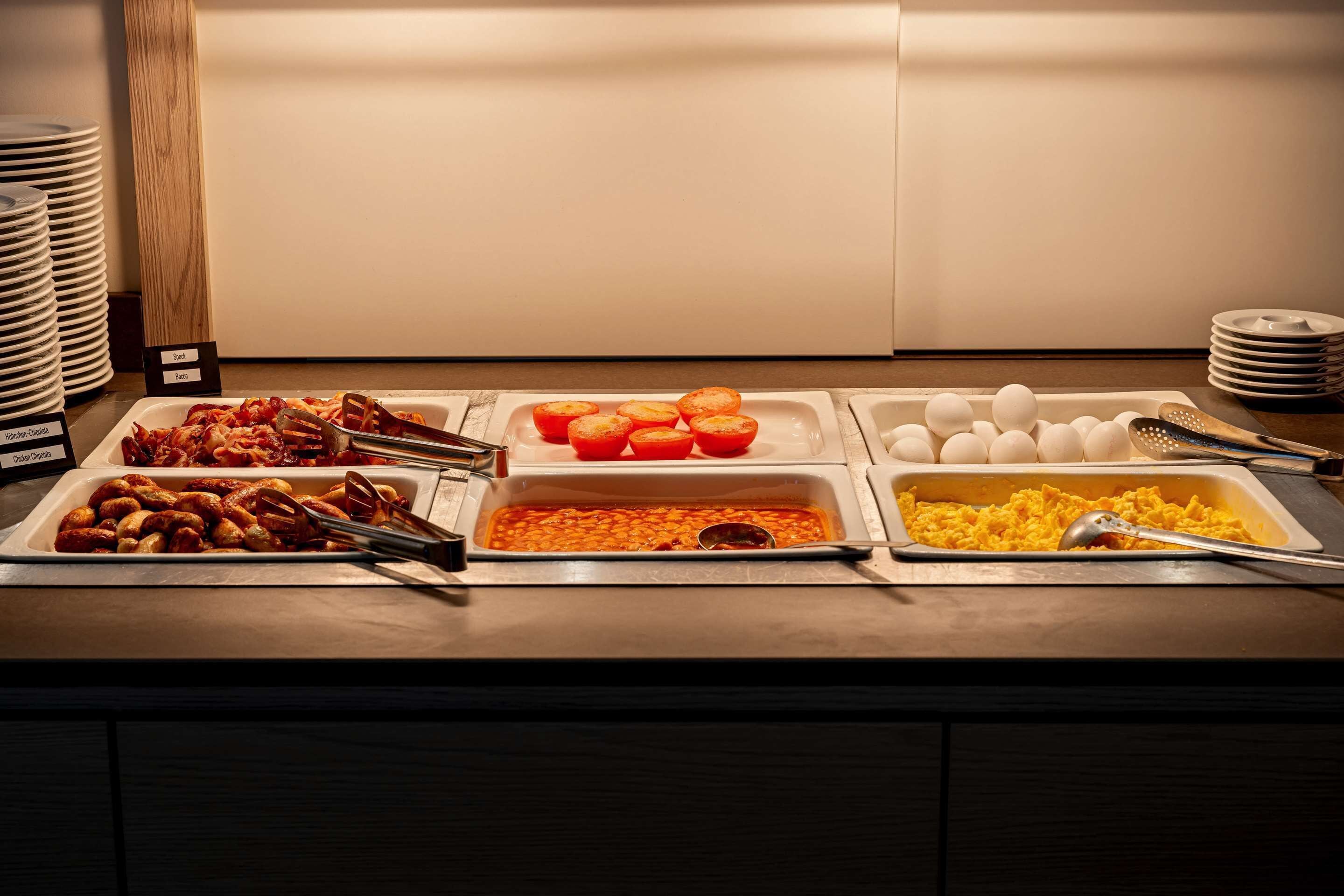 Buffet com áreas de comida identificadas, incluindo salsichas, tomates, ovos mexidos e feijão cozido.