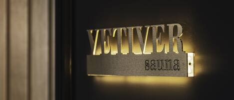 Uma placa metálica com o nome 'VETIVER' e a palavra 'Sana' sobre um fundo escuro.