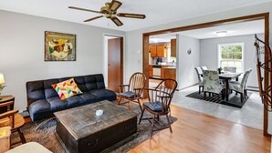 Uma sala de estar com sofá, mesa de centro e cadeiras, um espaço de jantar com mesa e cadeiras, e uma cozinha visível através de uma porta aberta.