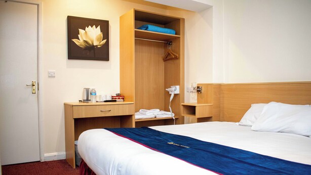 Quarto de hotel com cama, guarda-roupa de madeira, porta-toalhas, obra de arte na parede e uma pequena mesa com cafeteira.
