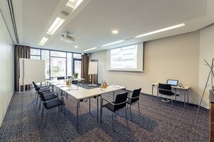 Uma sala de conferência com uma mesa comprida, cadeiras pretas, uma tela de projetor e um laptop em uma mesa.