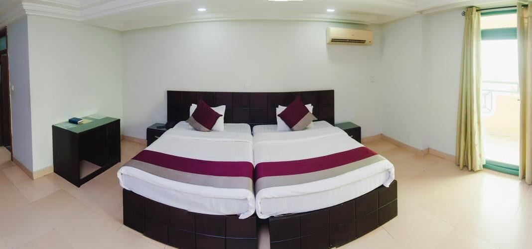 Cama de casal com cabeceira escura, duas mesas de cabeceira, uma escrivaninha pequena e uma janela com cortinas.